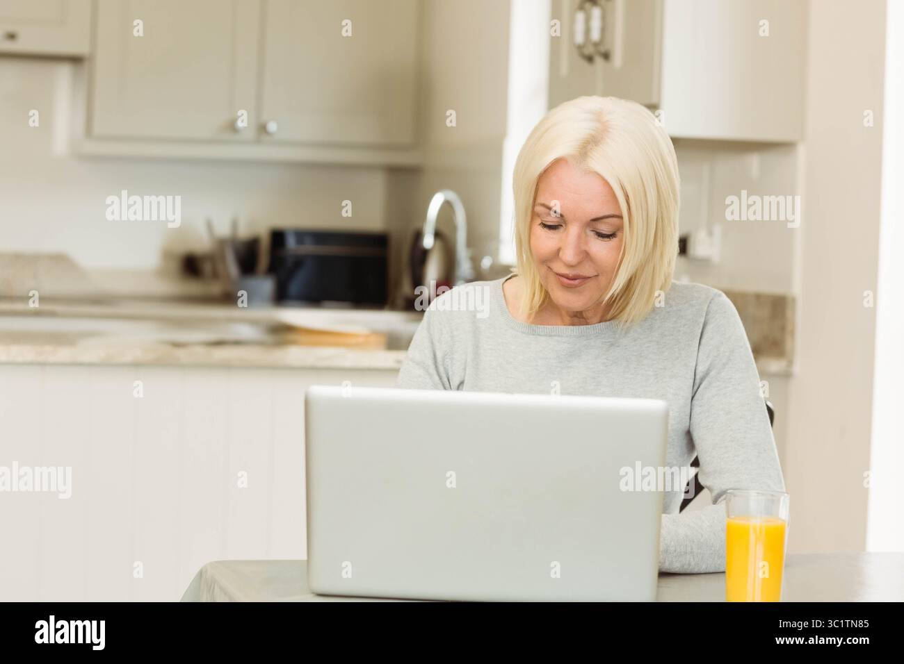 Woman browsing on laptop in modern kitchen while sipping orange juice ...