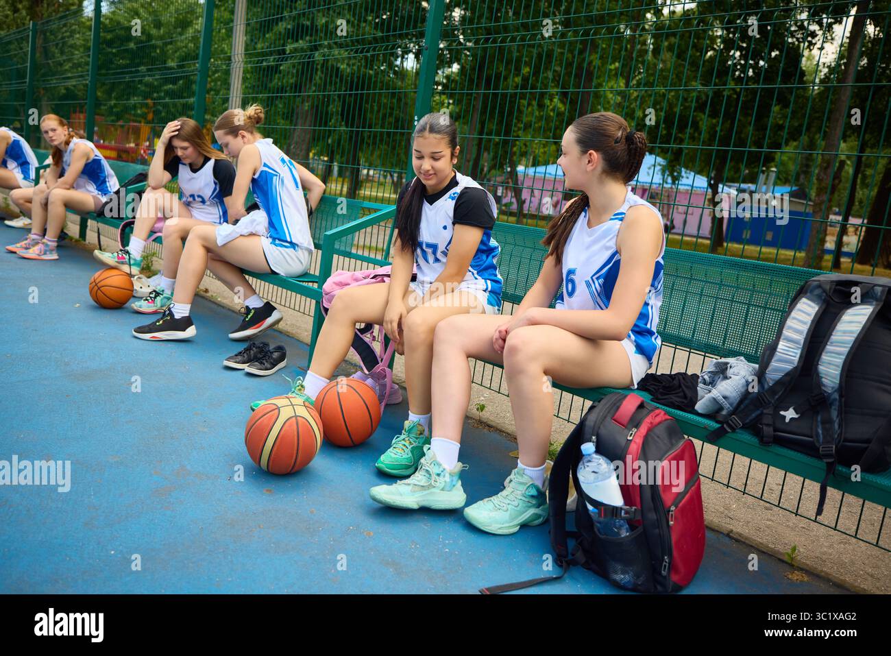 Female basketball athletes sitting on bench in team uniforms at outdoor ...