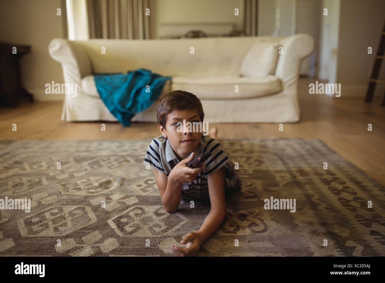 Boy lying on patterned rug in living room holding remote control and ...