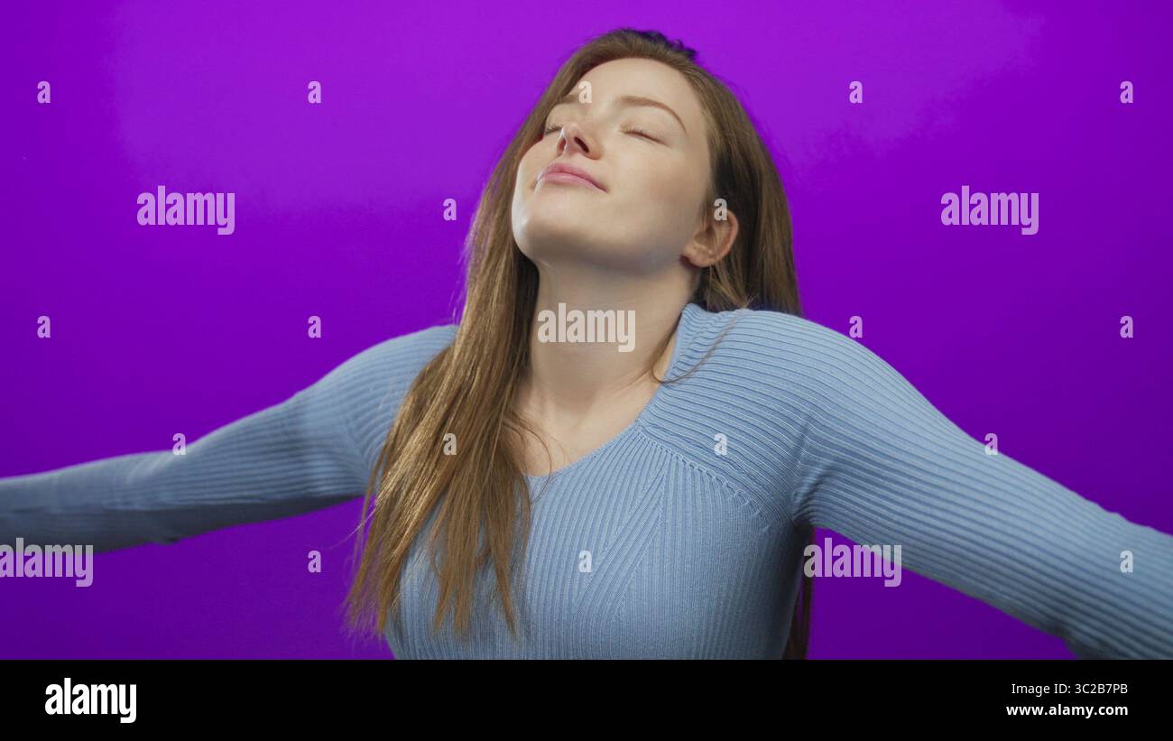 Woman stretches arms wide with lifted chin and closed eyes in vibrant purple studio; serenity ...