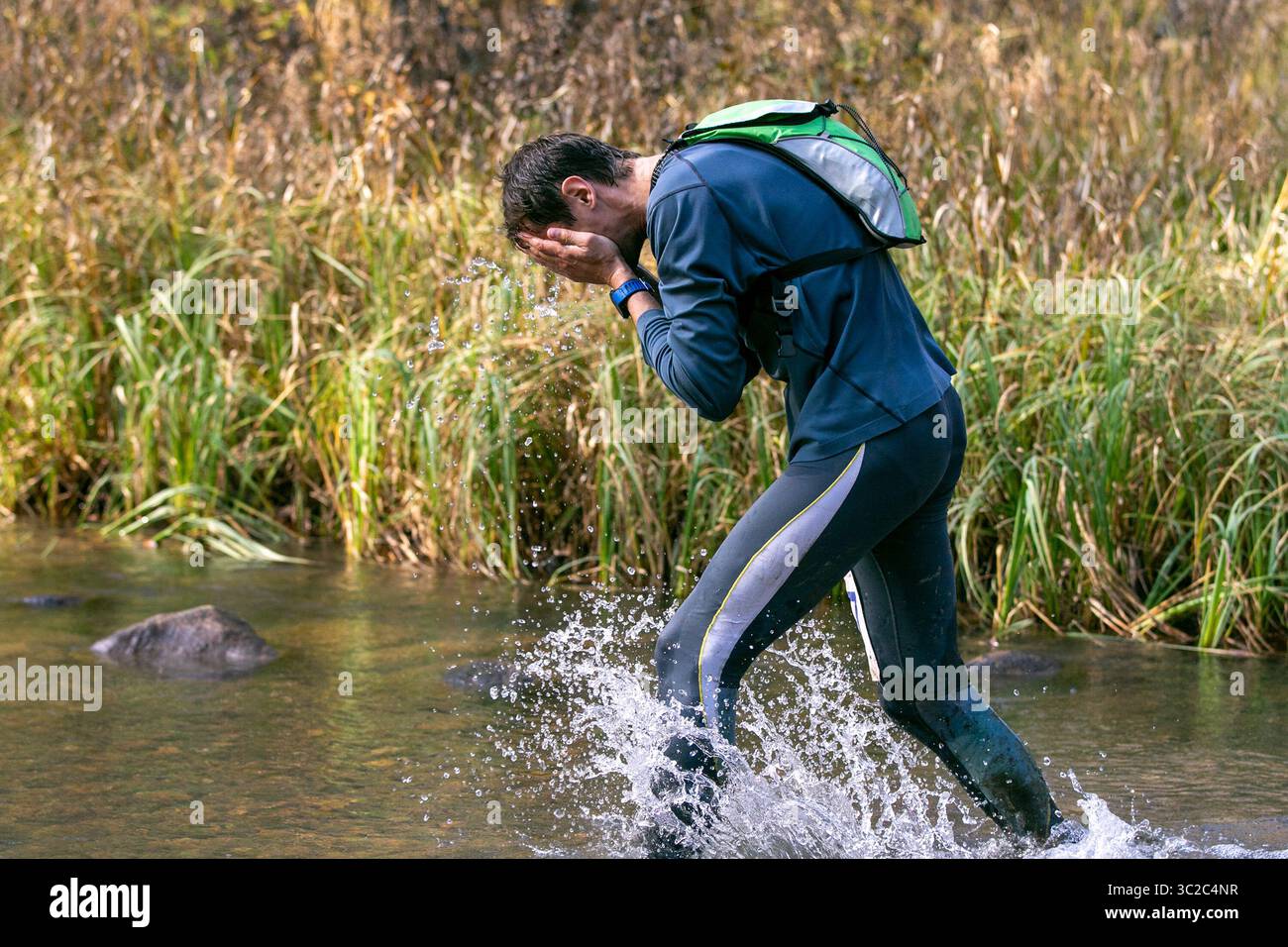 male runner wading through river refreshes his face with water during ...