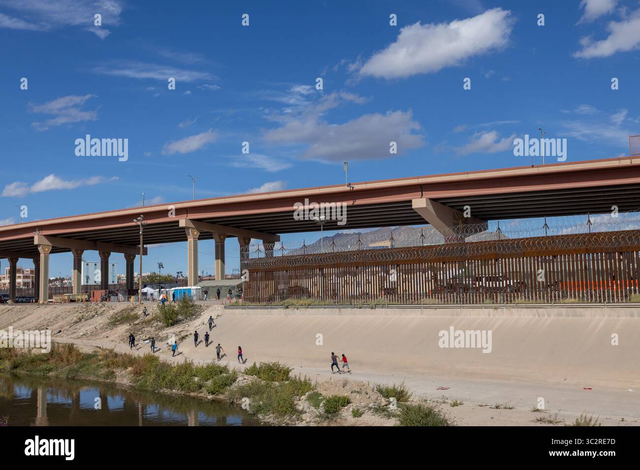 People walk near a border fence under a highway in the desert near ...