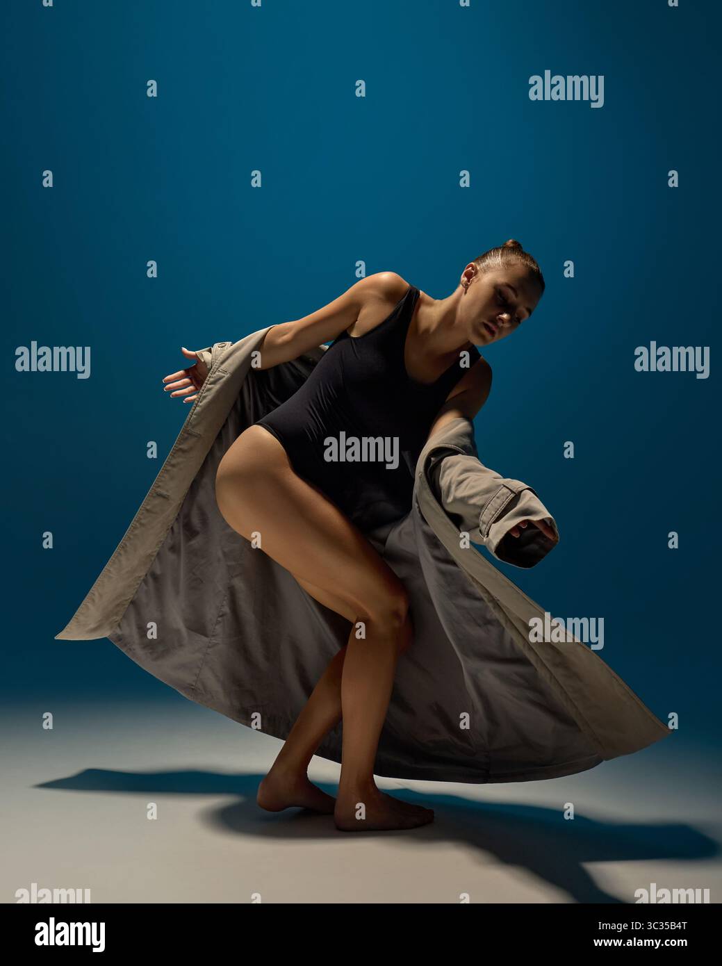 Young woman bending in dramatic spotlight with trench coat and ...