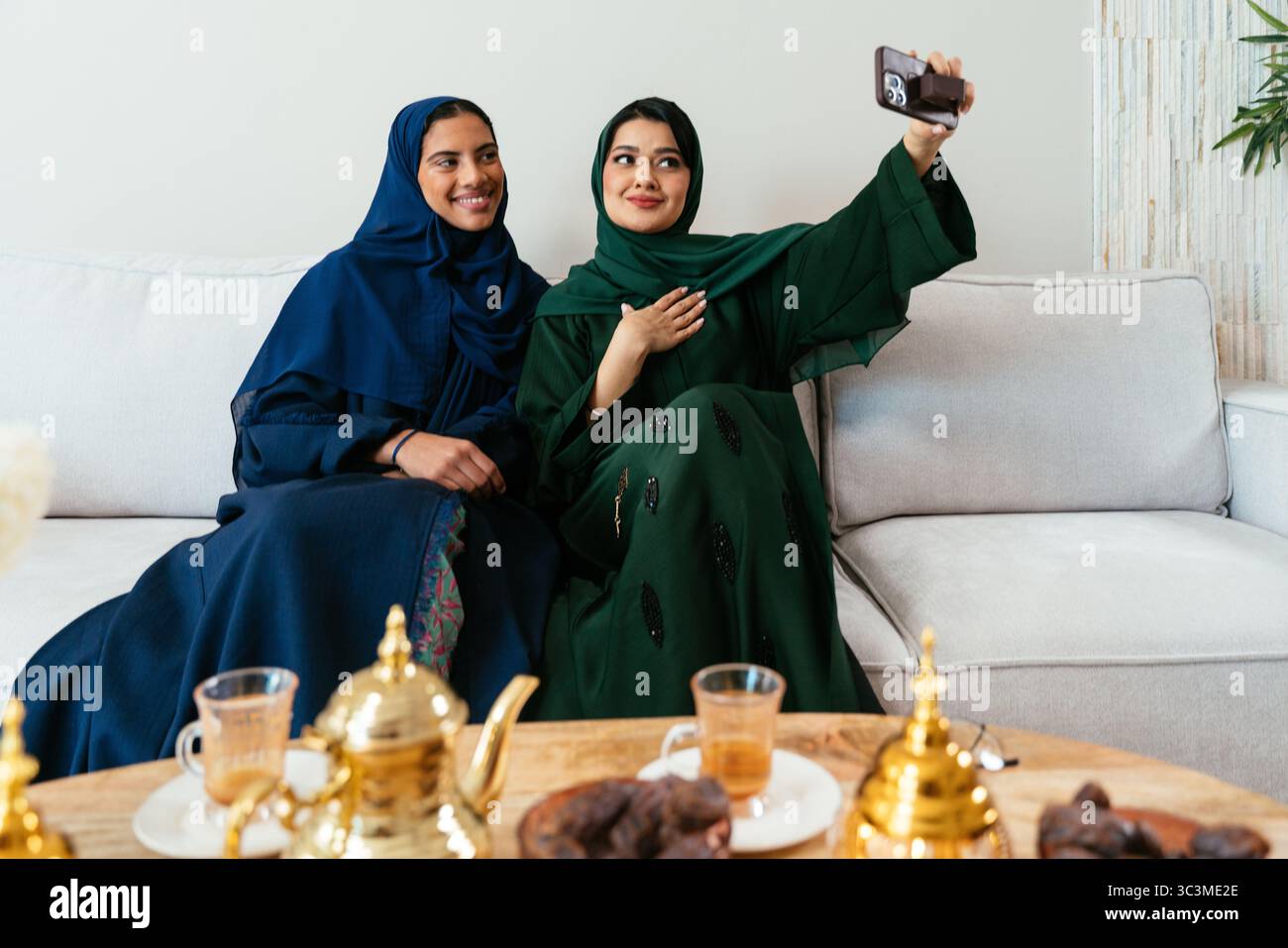 Two emirati women in traditional abayas sitting on a sofa, enjoying tea ...