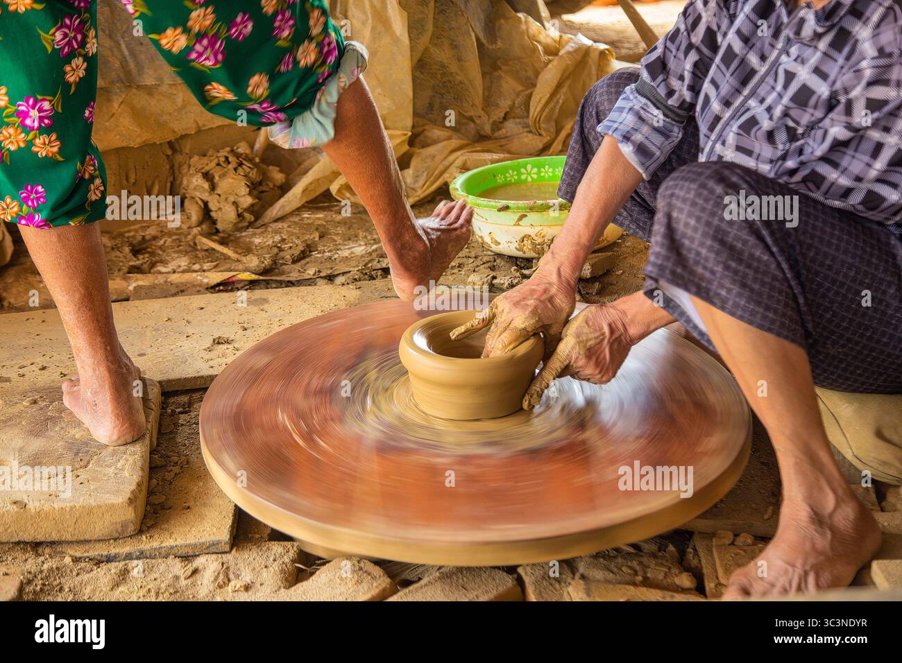 Two women work on a pottery wheel, one pedaling the wheel, the other ...