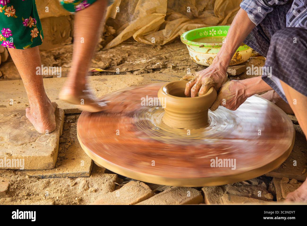 Two women work on a pottery wheel, one pedaling the wheel, the other ...
