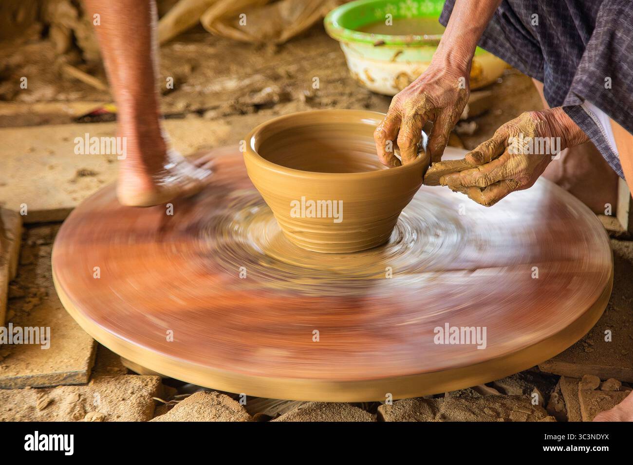 Two women work on a pottery wheel, one pedaling the wheel, the other ...
