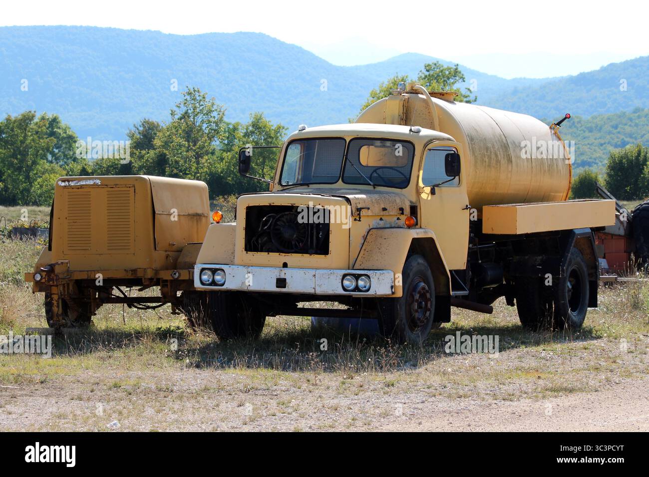 A vintage yellow water tanker truck, missing its front grille and ...