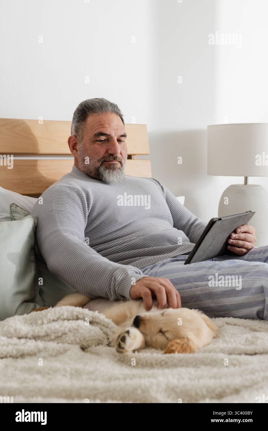 Mature man holding tablet and petting golden retriever puppy while ...