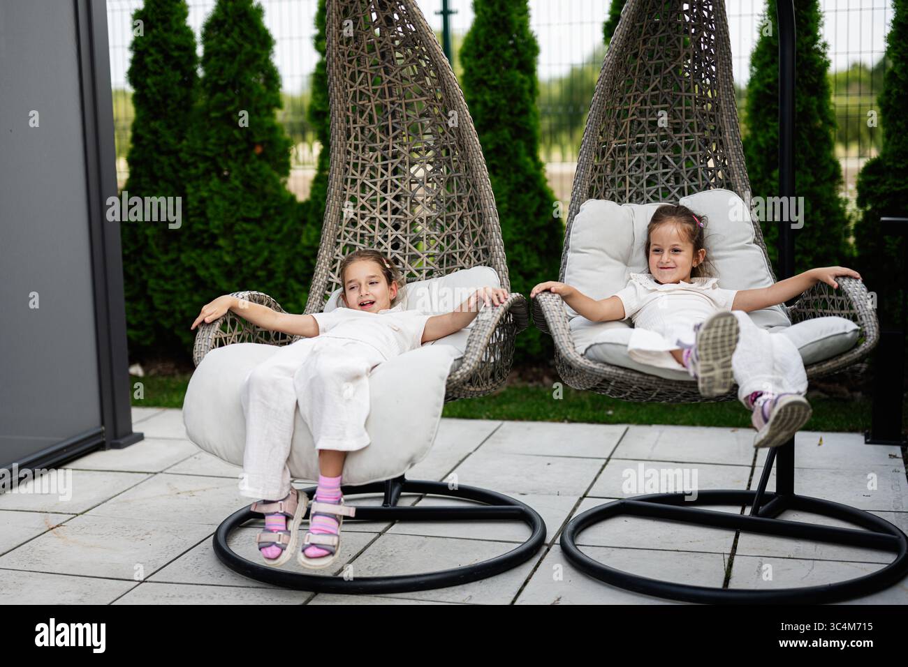 Two children sitting on comfortable patio chairs in a green garden area ...