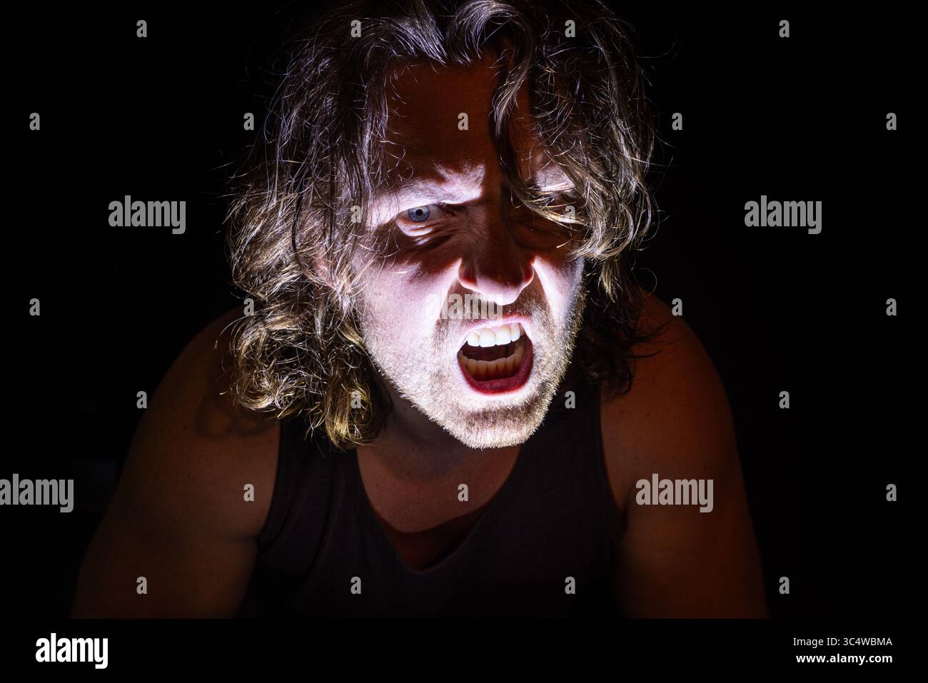 Man with long scraggly hair holding a flashlight under his shin in a ...