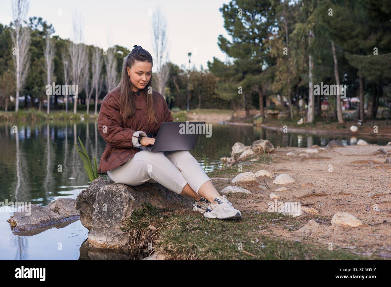 A young Hispanic woman with long dark hair sits on a rock by a lake ...