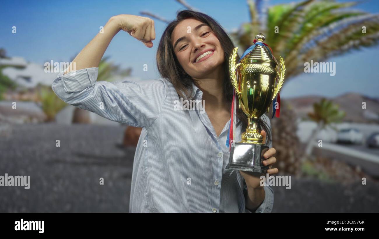 Woman holds trophy and flexes arm on city street with palm trees and ...