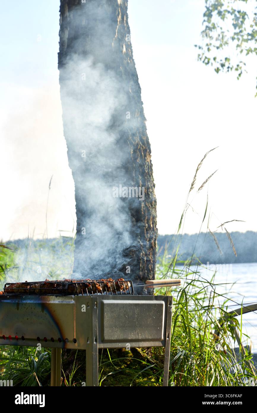 Smoky barbecue grill with meat cooking outdoors near the river. Summer ...
