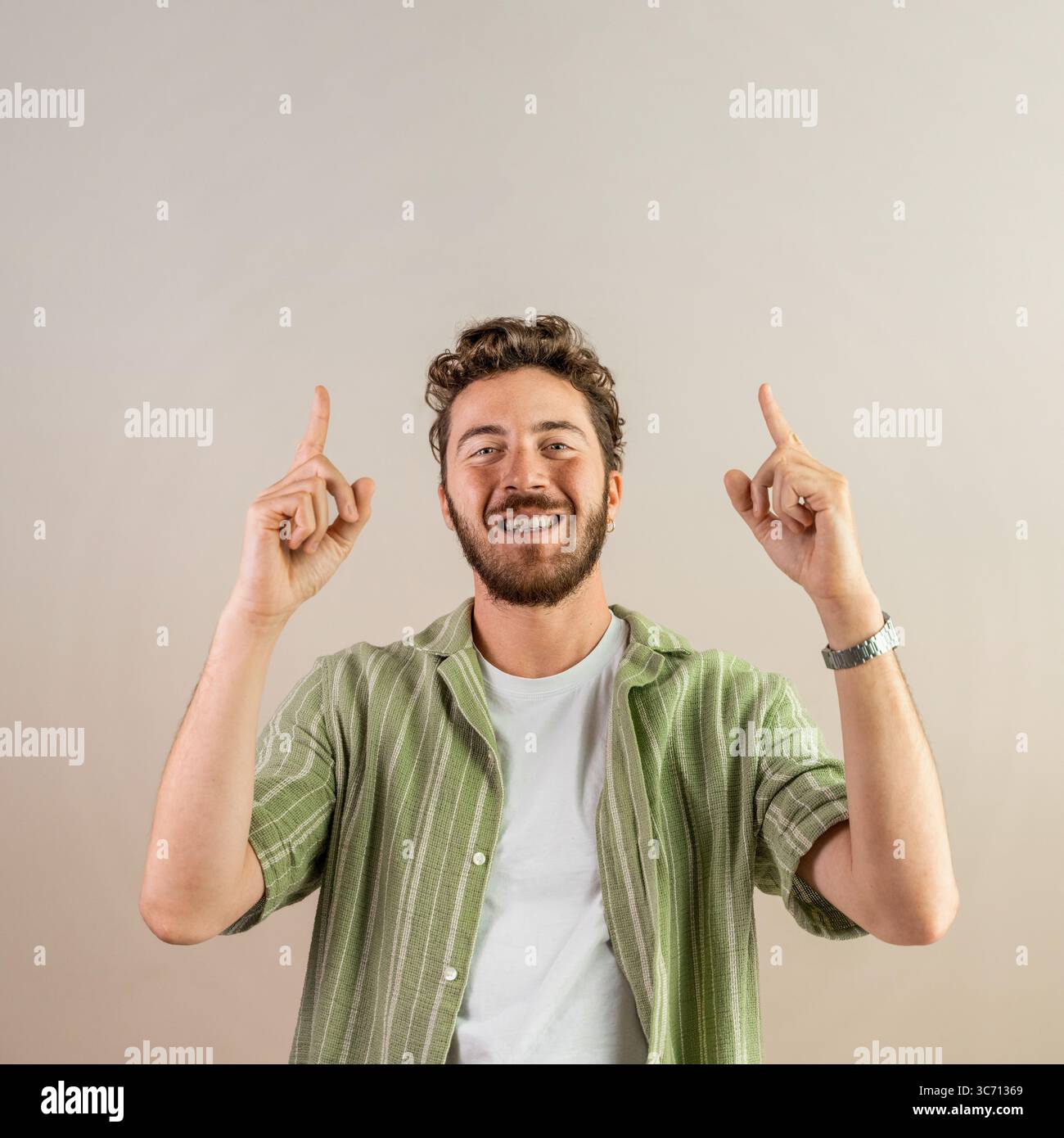 A cheerful smiling young man with a beard looks at the camera and points upwards with both hands ...
