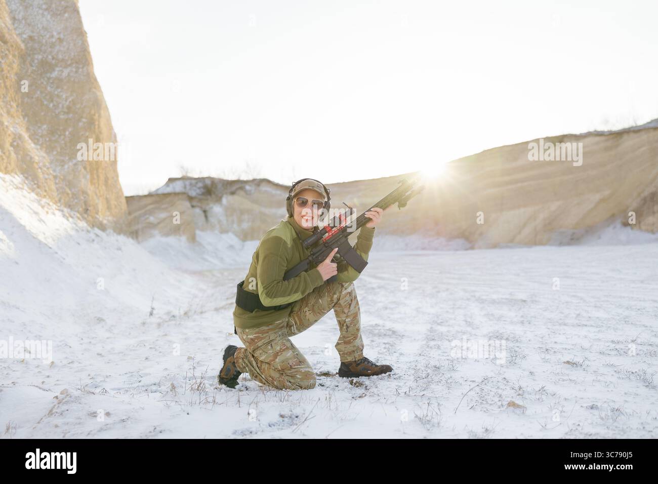 Female soldier kneeling and aiming with a rifle in a snowy shooting range during a winter ...