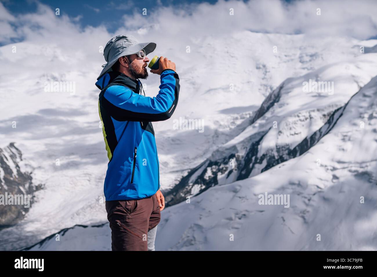 Climber in a hat drinking hot tea on Yuhin peak during Lenin peak ascent under mountain peaks in ...