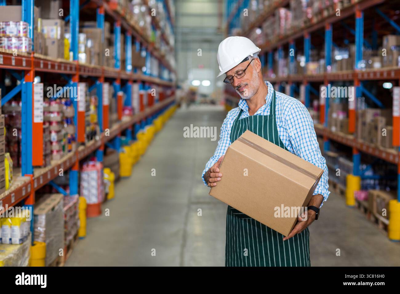 Mature adult warehouse worker inspecting cardboard box in aisle with ...
