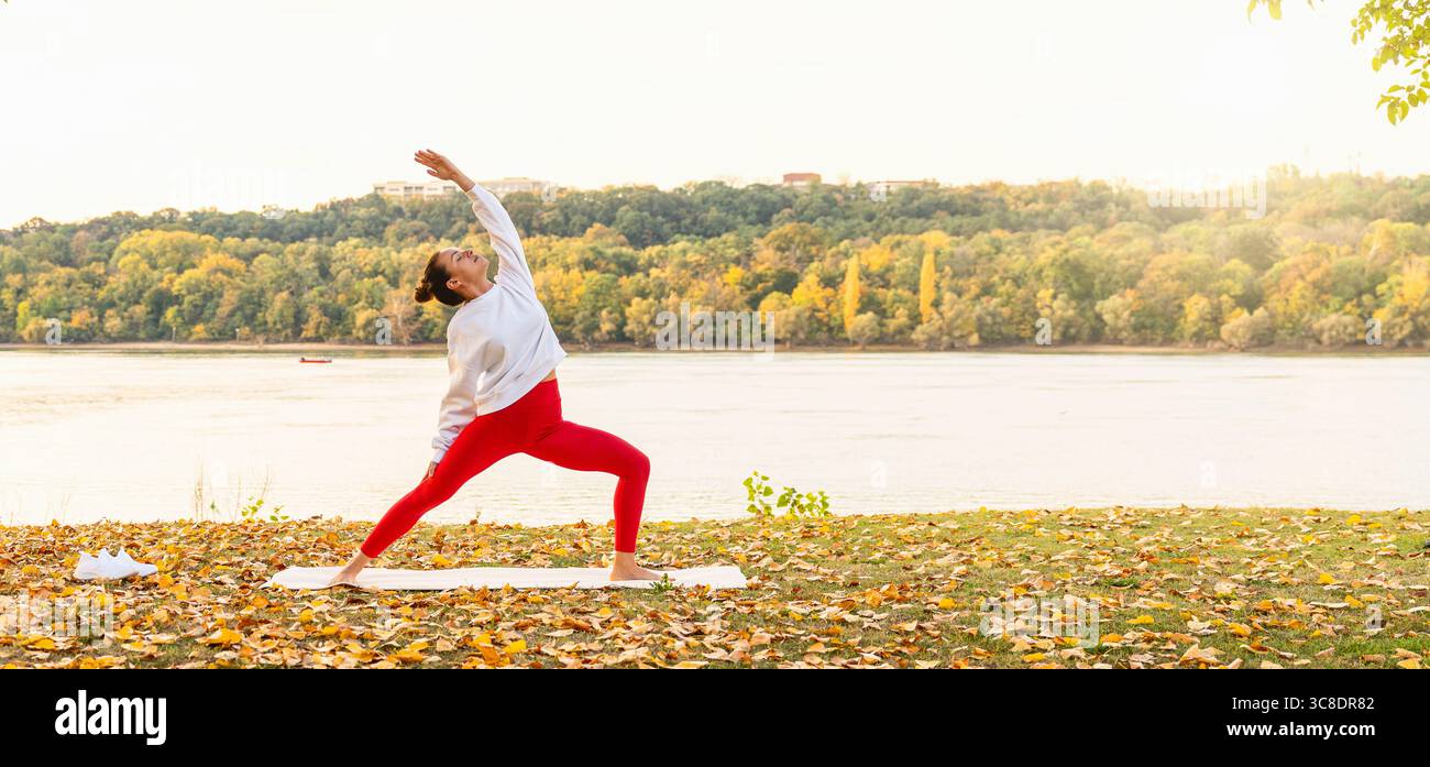Woman doing outdoor yoga exercise by riverside during fall season ...