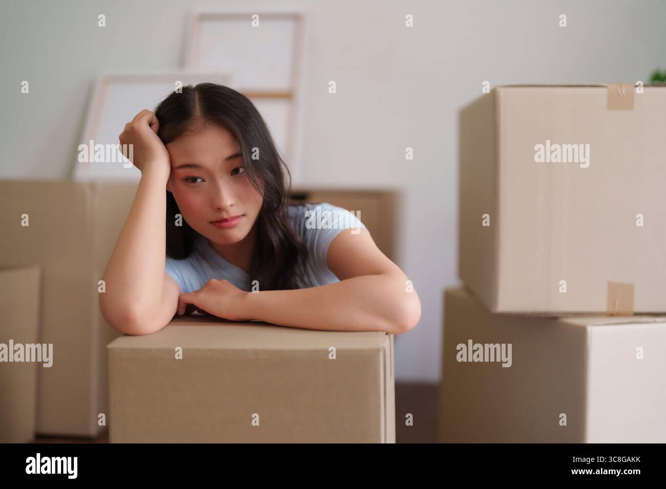 Young Asian female leans on moving box feeling overwhelmed with ...