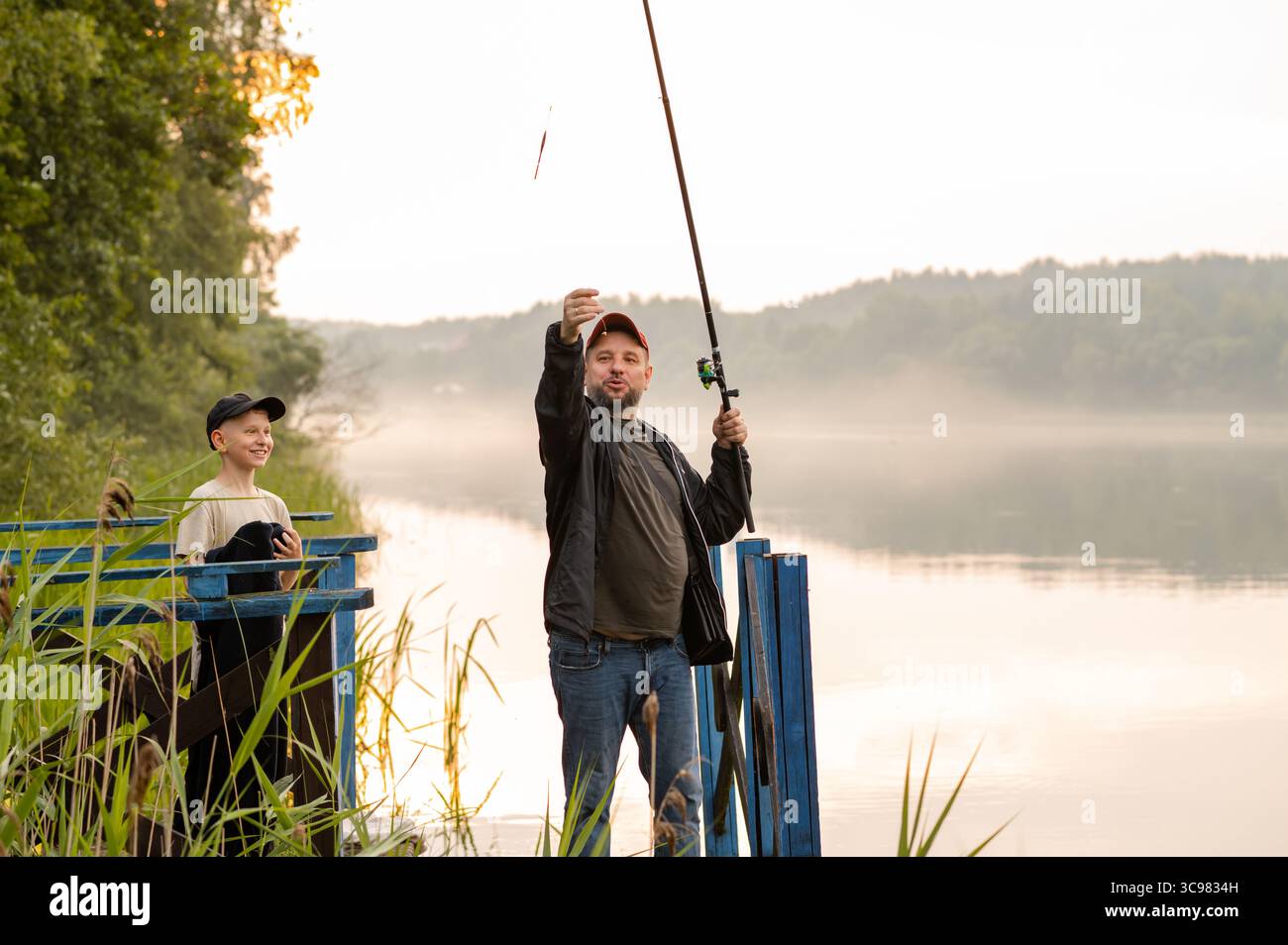 Father helping his son prepare fishing gear on a misty morning by the ...