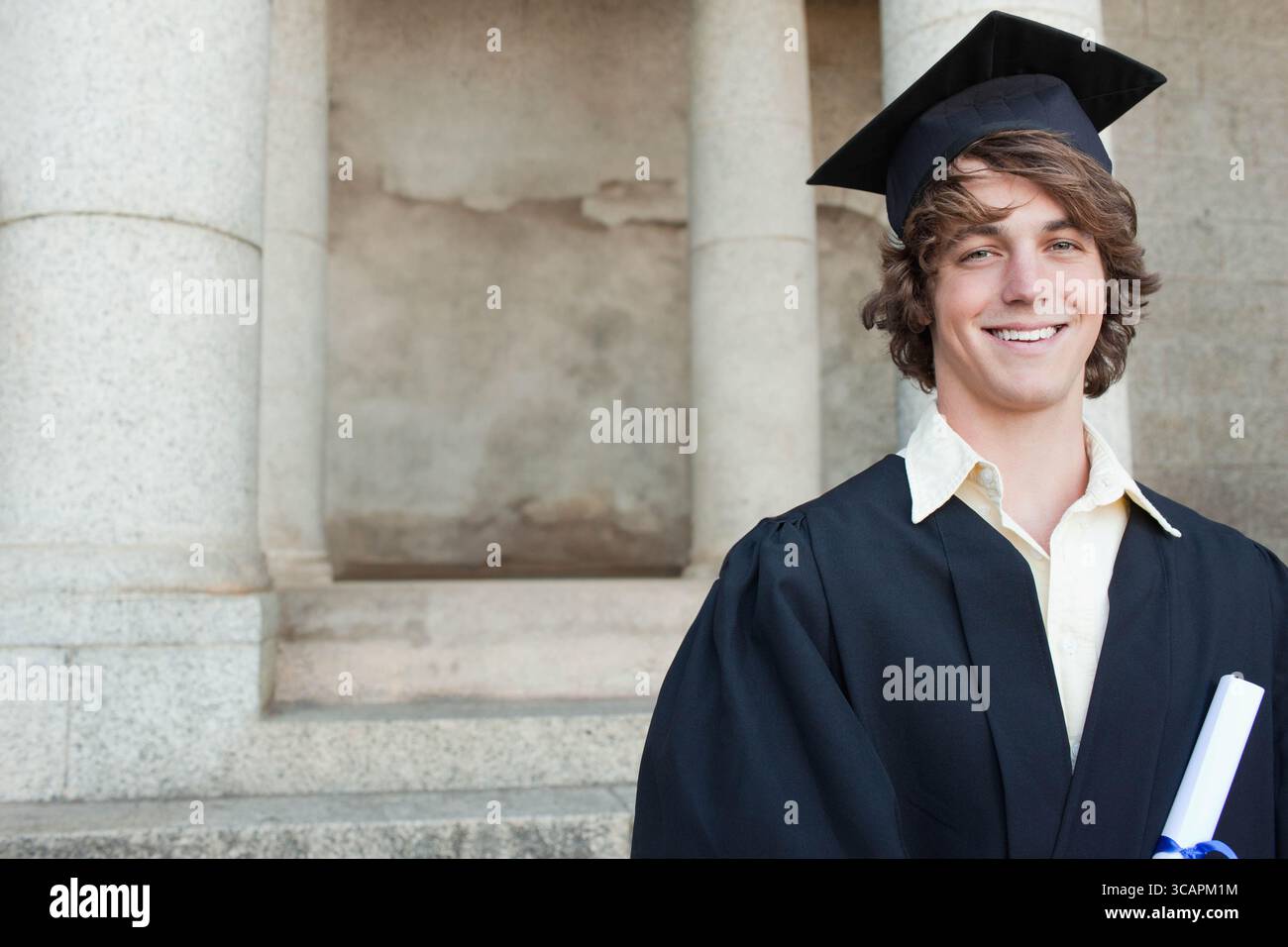 Male graduate holding diploma scroll standing by steps and columns in ...