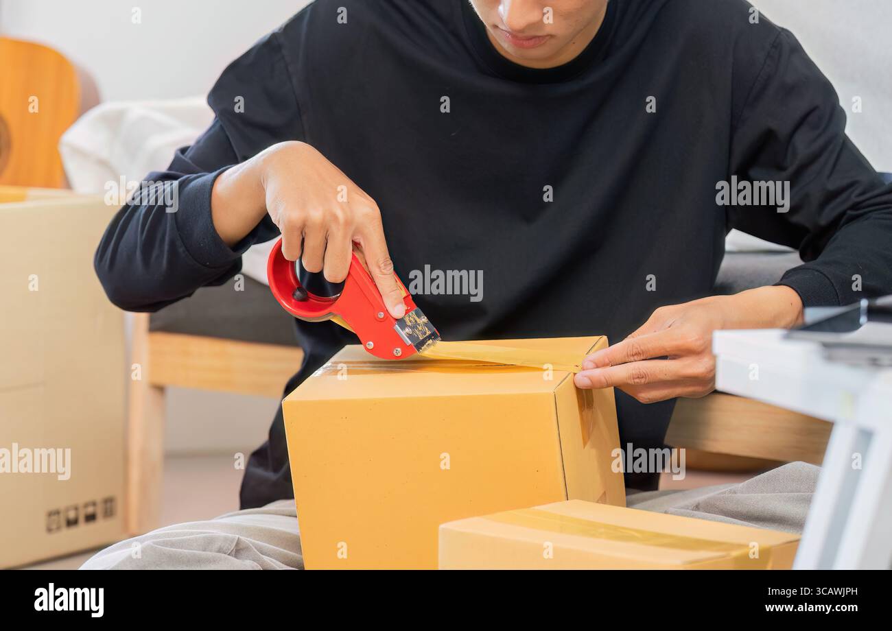 Unpacking. Young man cutting open a cardboard box with a tape cutter ...