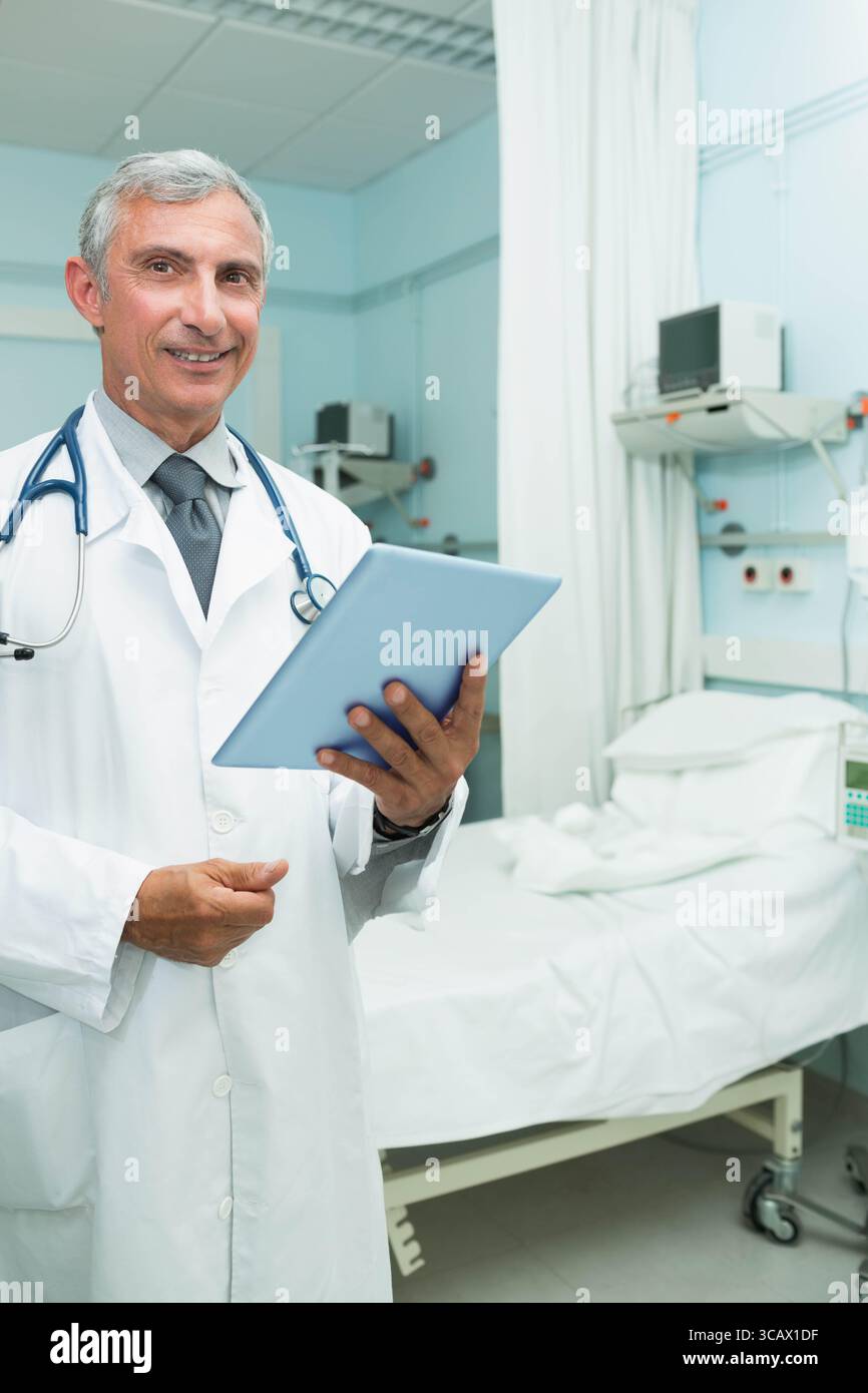 Senior male doctor standing at bedside holding tablet wearing lab coat ...