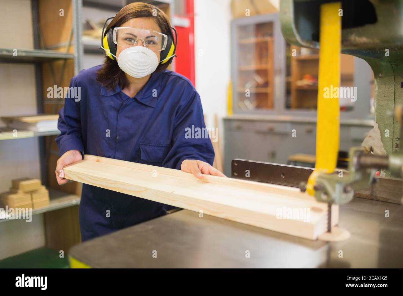 Female woodworker guiding wooden plank through powered bandsaw wearing ...
