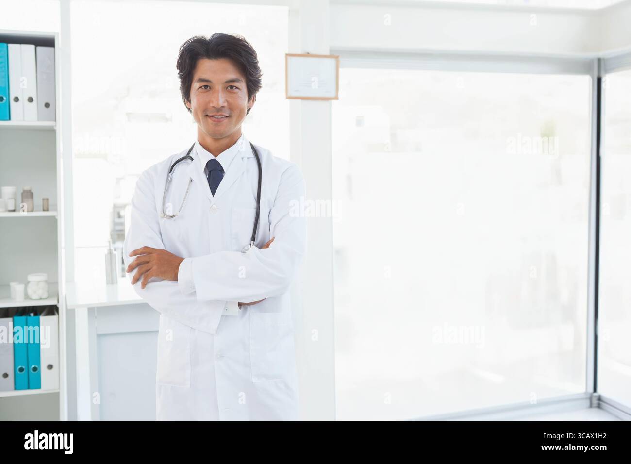 Japanese male doctor standing with arms folded by clinic shelving ...