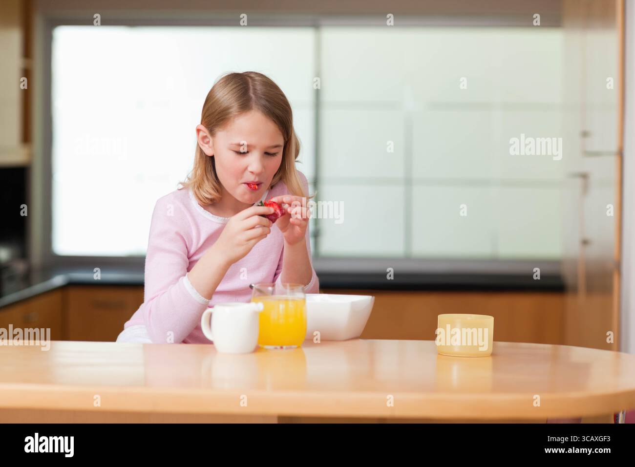 School-age girl sitting on stool and eating ripe strawberry at wooden ...
