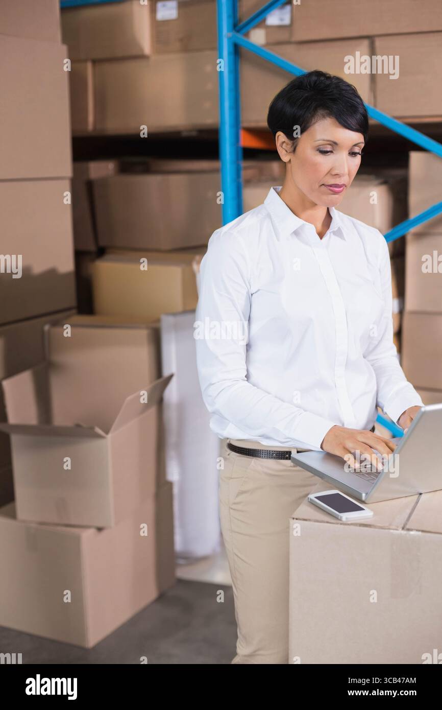Female warehouse clerk leaning on cardboard boxes checking laptop and ...