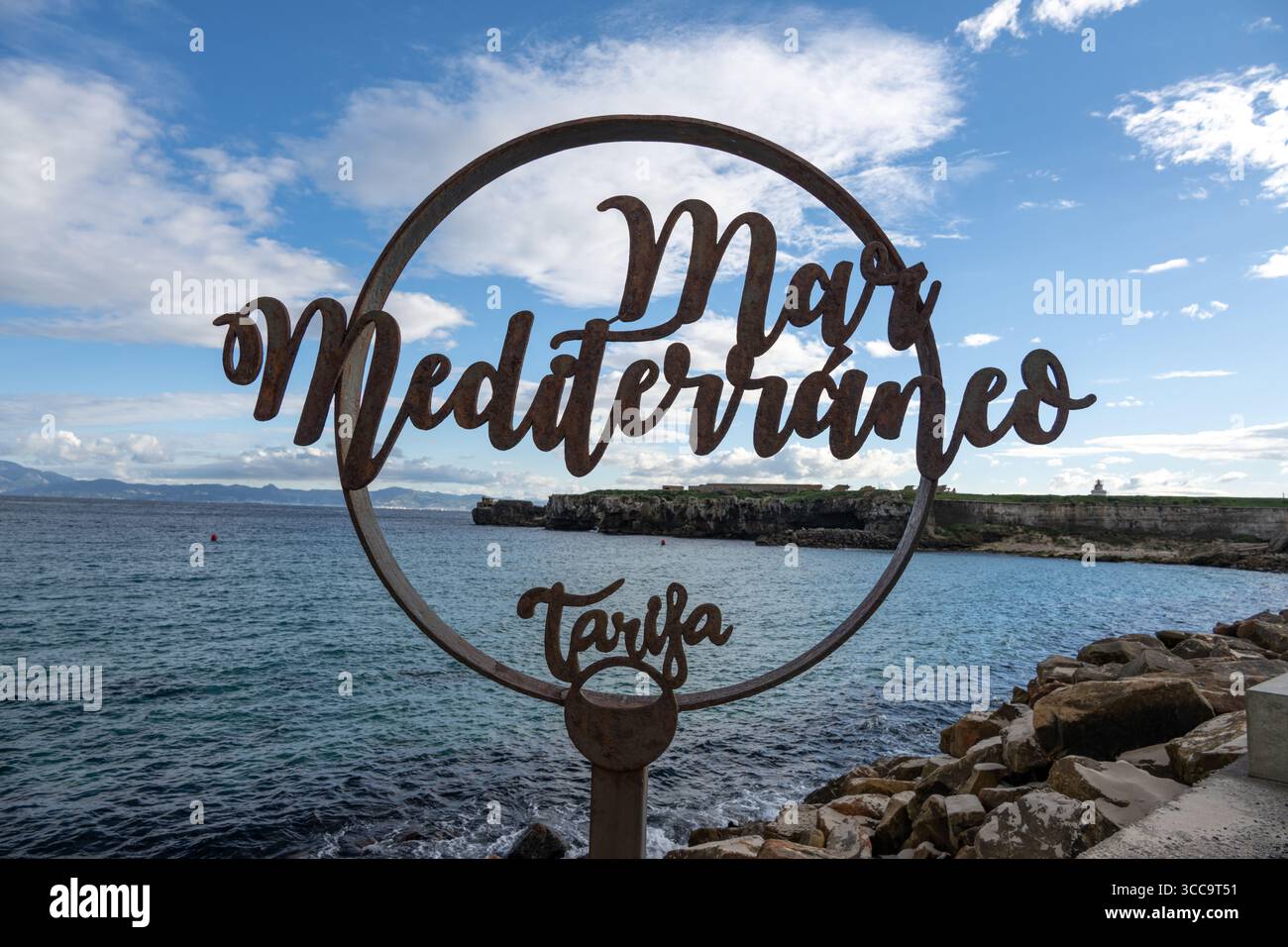 A metal sign with 'Mar Mediterráneo Tarifa' stands against a scenic ...