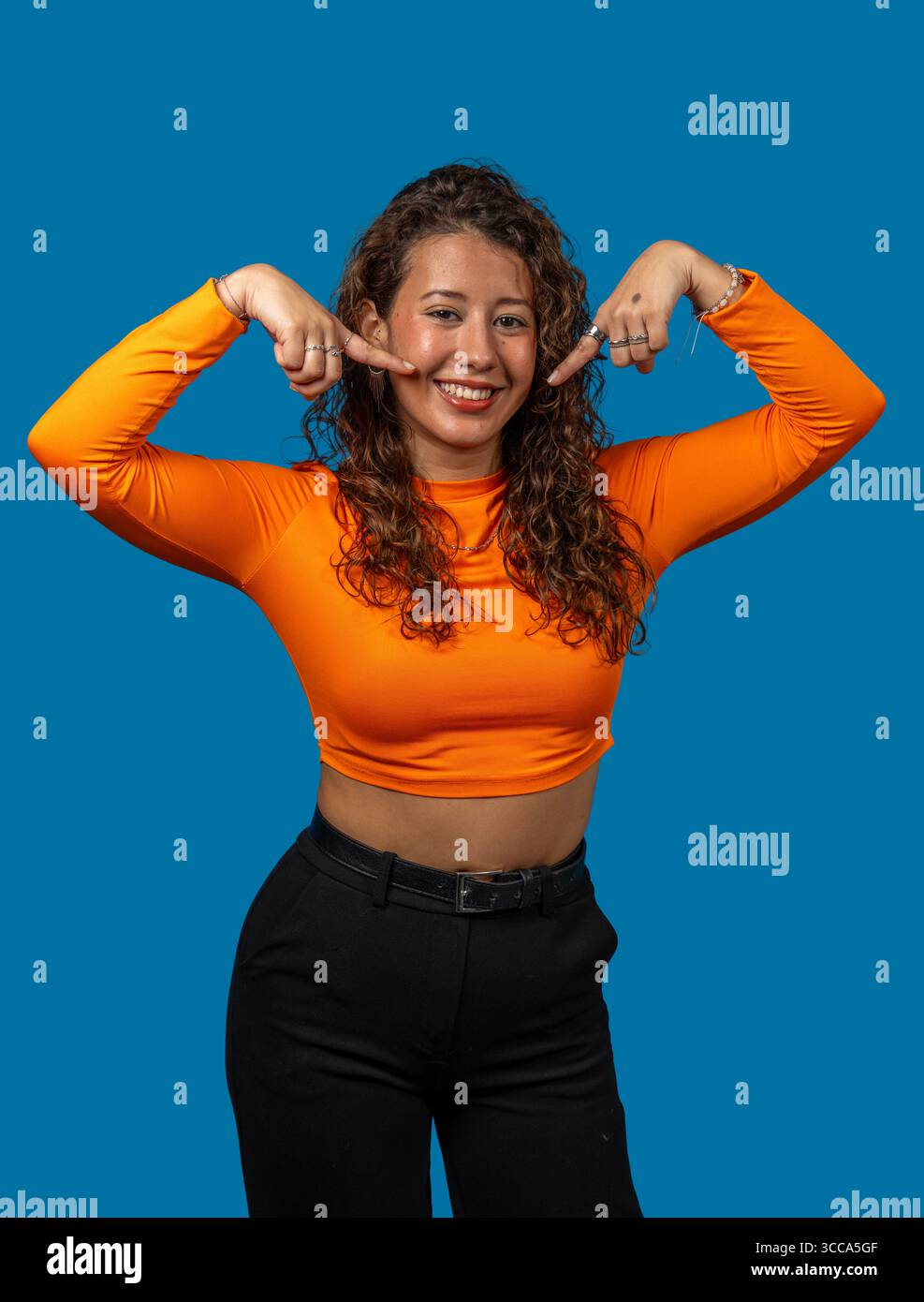 Studio portrait of cheerful young woman pointing at her smile against a ...