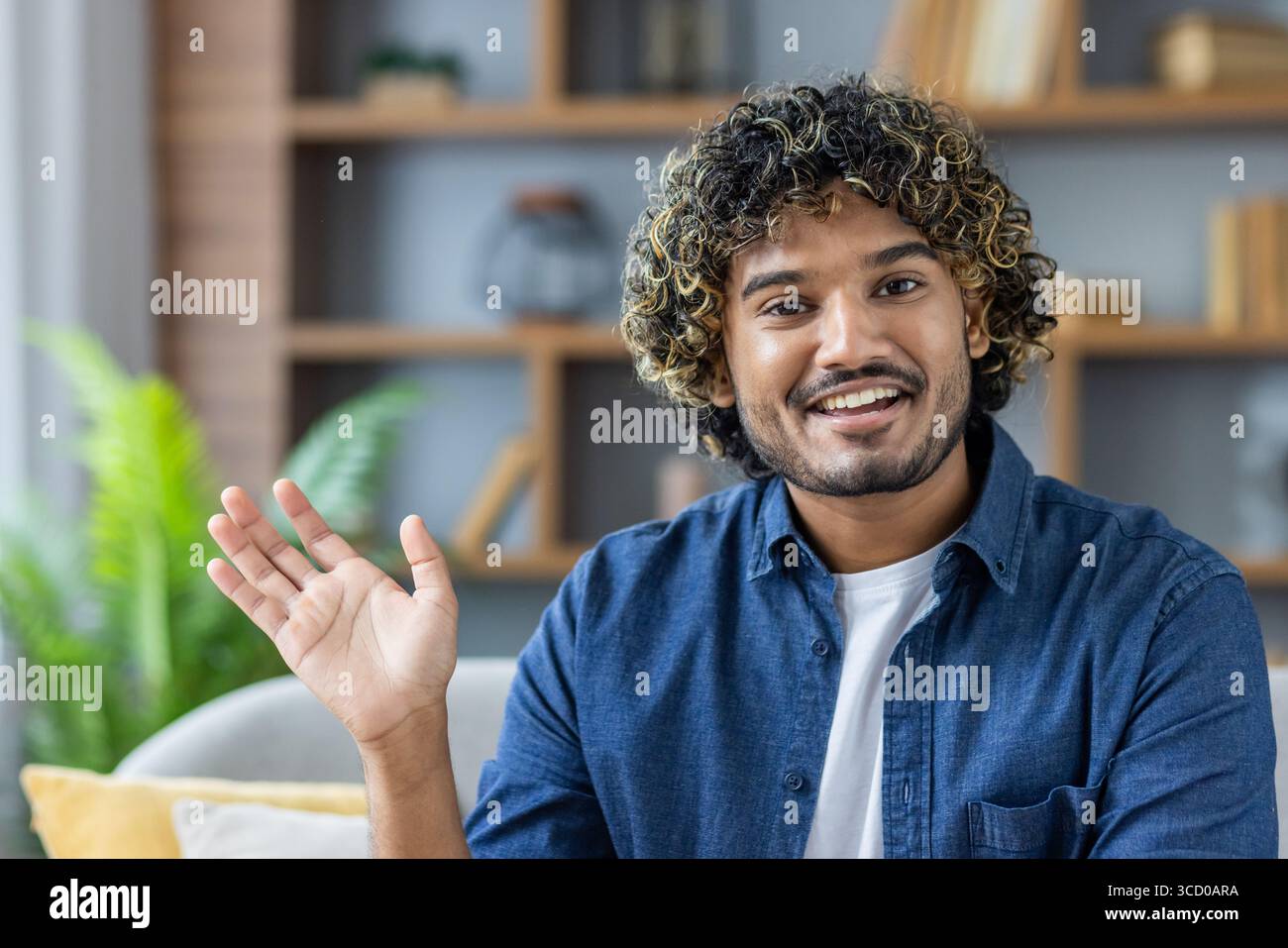A cheerful young man smiles and waves, seated in a cozy, modern home ...