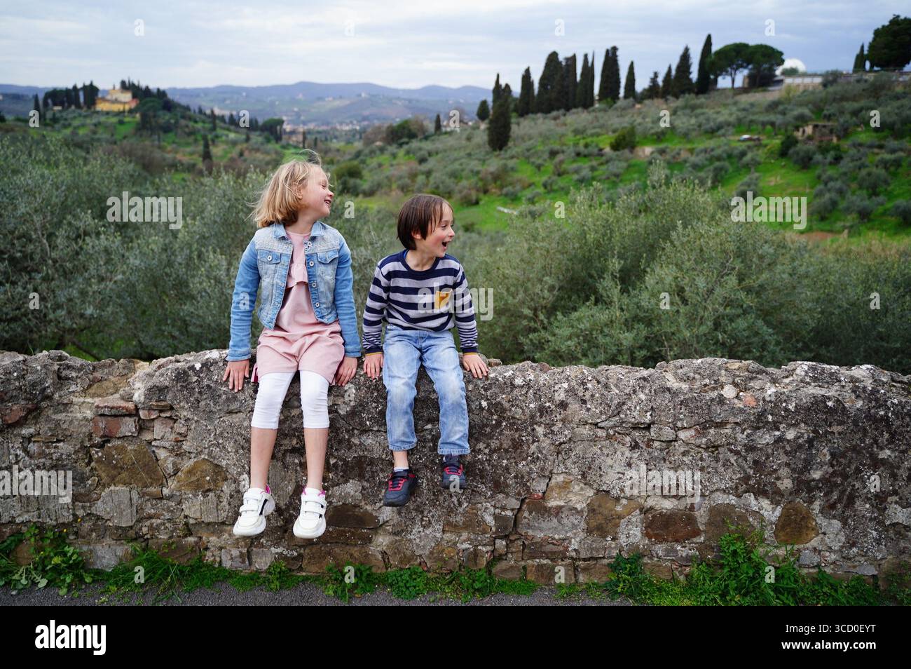Two children sit on a stone wall, laughing together against a scenic ...