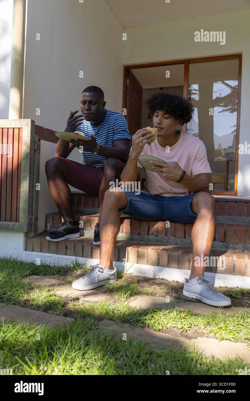 Diverse male friends biting into burgers sitting on brick steps on ...