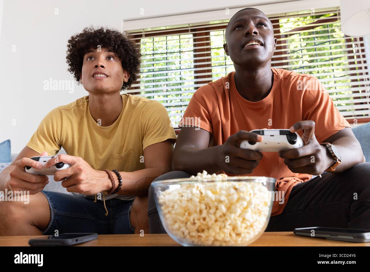 Diverse male friends holding wireless controllers and eating popcorn on ...
