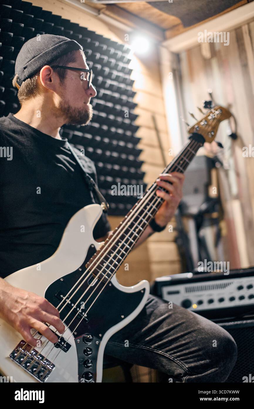 Young male bassist with glasses and cap focused on playing bass guitar ...