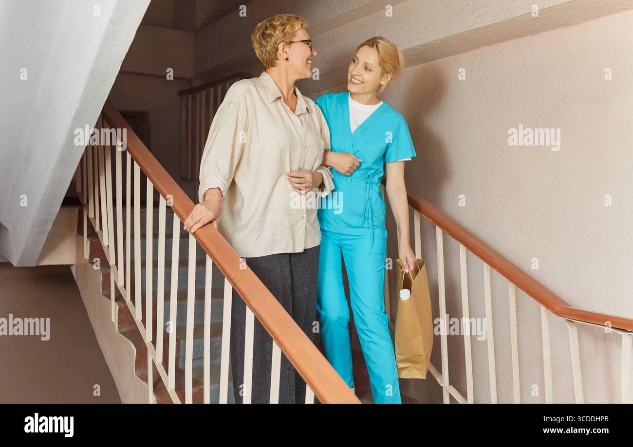 Young female caregiver helping to senior woman on stairs with grocery ...
