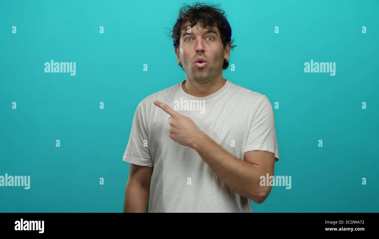 Hispanic man points aside surprised against blue background, wearing a ...