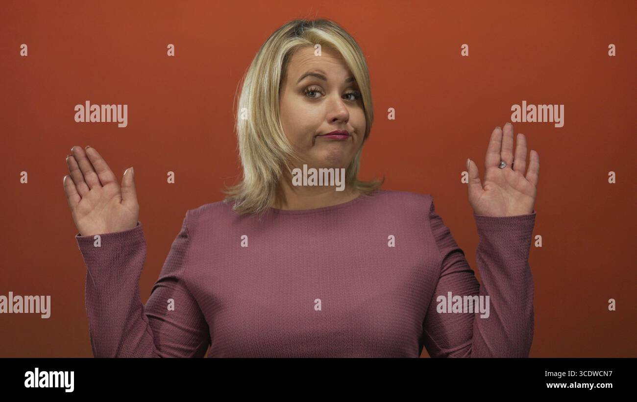 Young blonde woman holds up hands with palms forward in red studio with ...