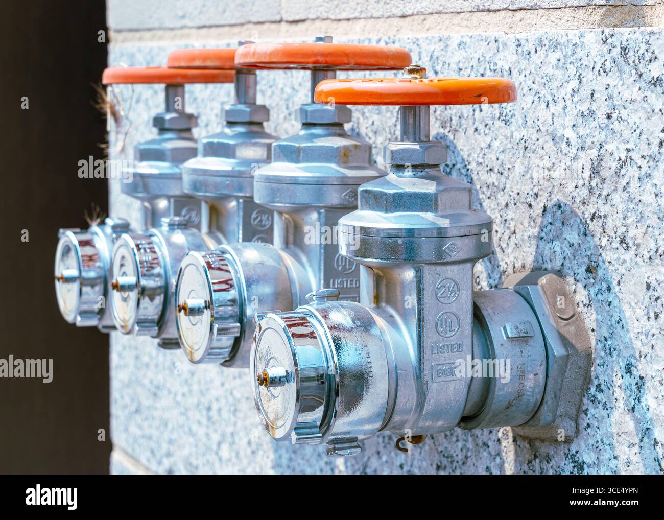 Four metallic fire hose valves with orange handles are mounted on a ...