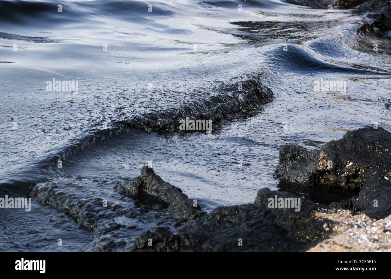 Rocky seashore hit by dark waves covered with rainbow sheen of oil ...