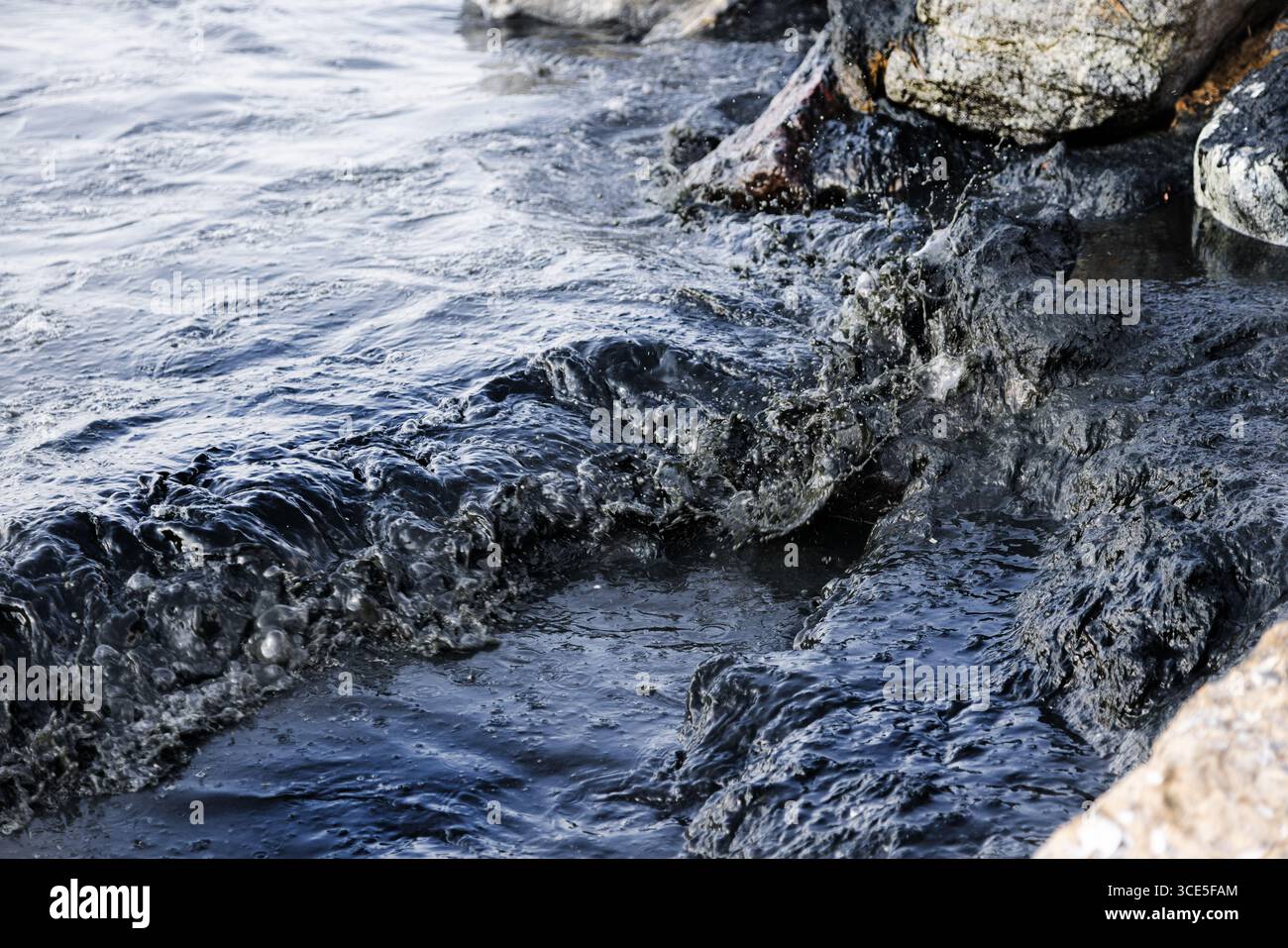 Rocky seashore hit by dark waves covered with rainbow sheen of oil ...