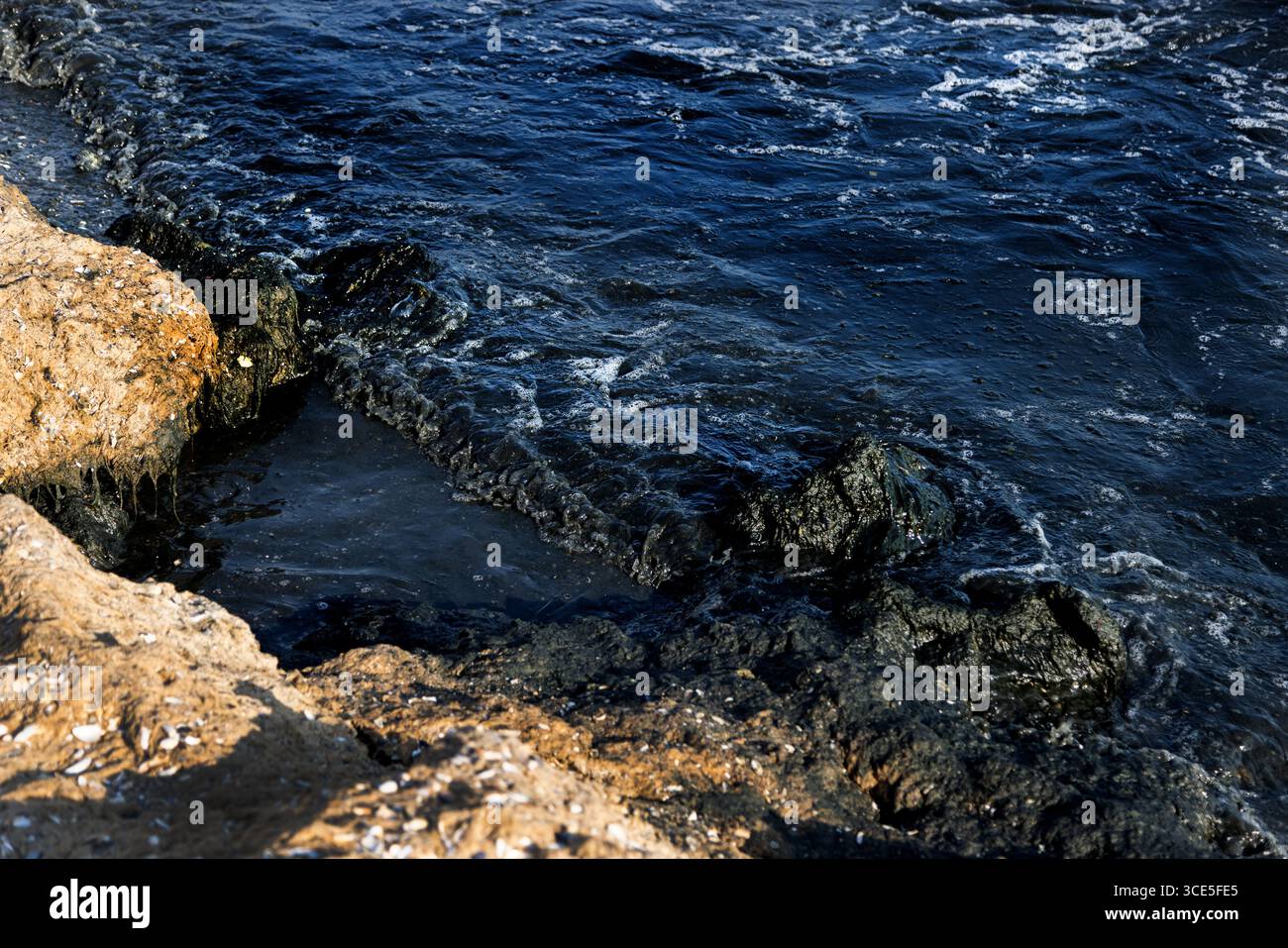 Rocky seashore hit by dark waves covered with rainbow sheen of oil ...