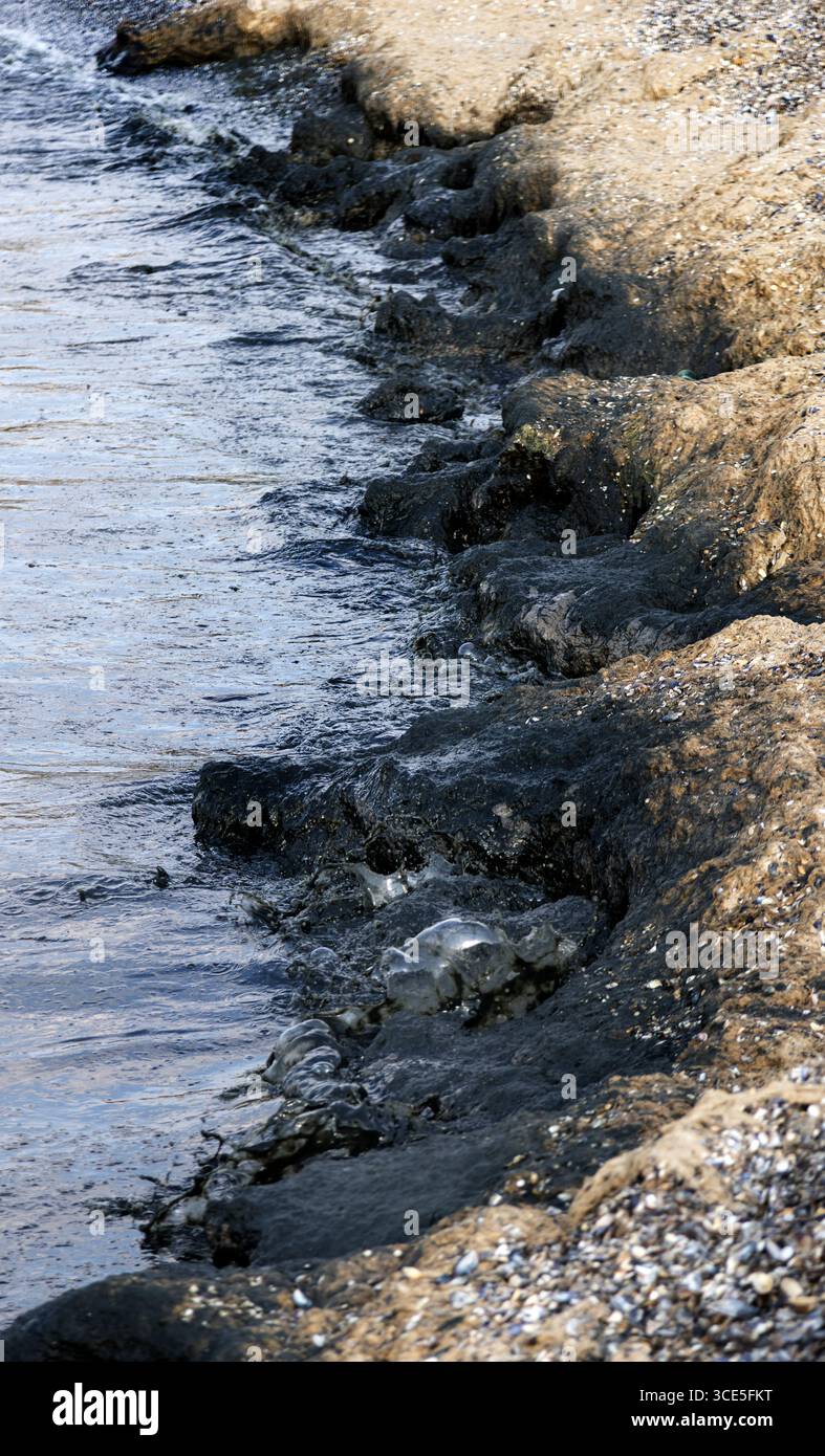 Rocky seashore hit by dark waves covered with rainbow sheen of oil ...