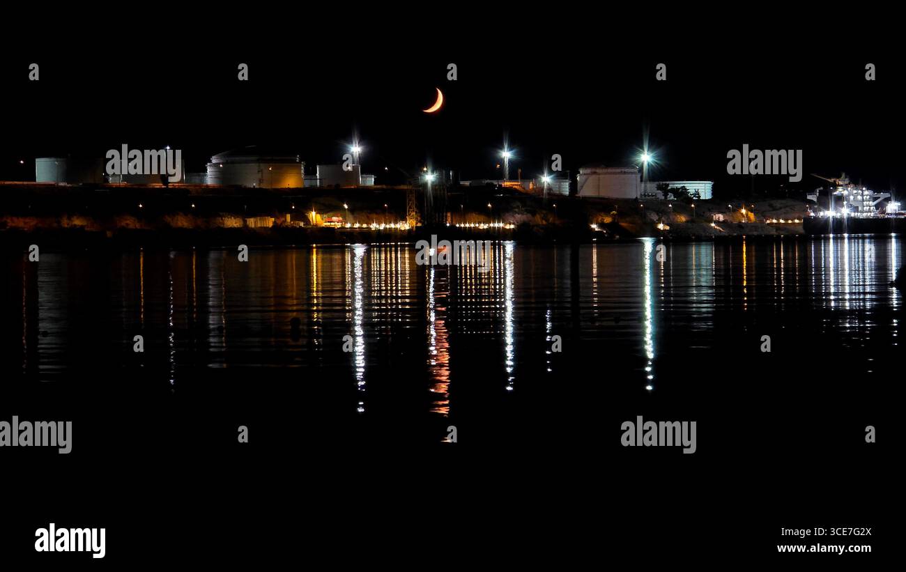 Crescent moon rises above illuminated oil tanks and waterfront terminal ...