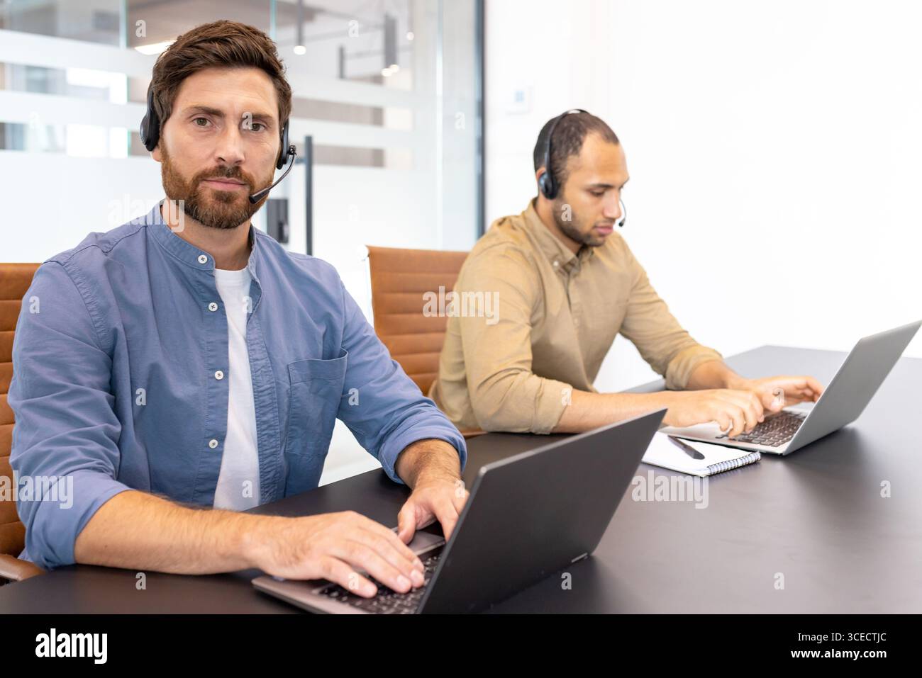 Two office professionals using laptops, wearing headsets, and engaged ...