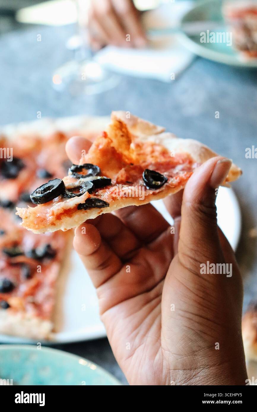 Hand holding a thin crust slice of pepperoni and olives pizza, another ...
